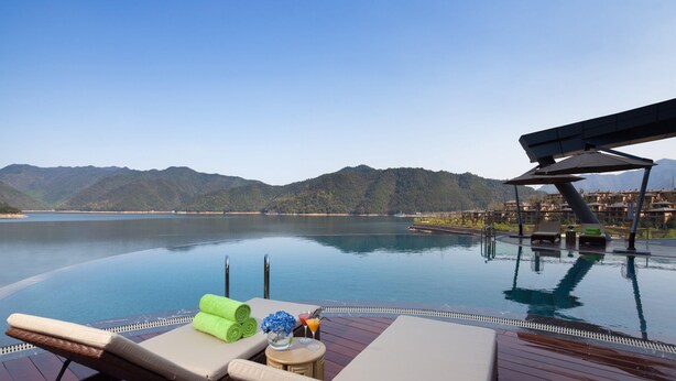 Uma piscina infinita com espreguiçadeiras, uma mesa com bebida e vista para as montanhas.