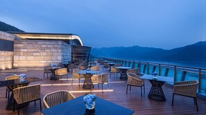Um terraço com mesas e cadeiras, com vista para um corpo de água e montanhas.