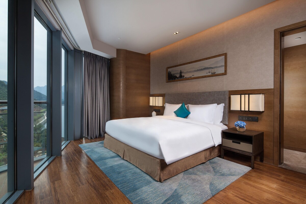 Quarto de hotel com uma cama grande, mesa de cabeceira, luminária na parede e janela com cortinas.