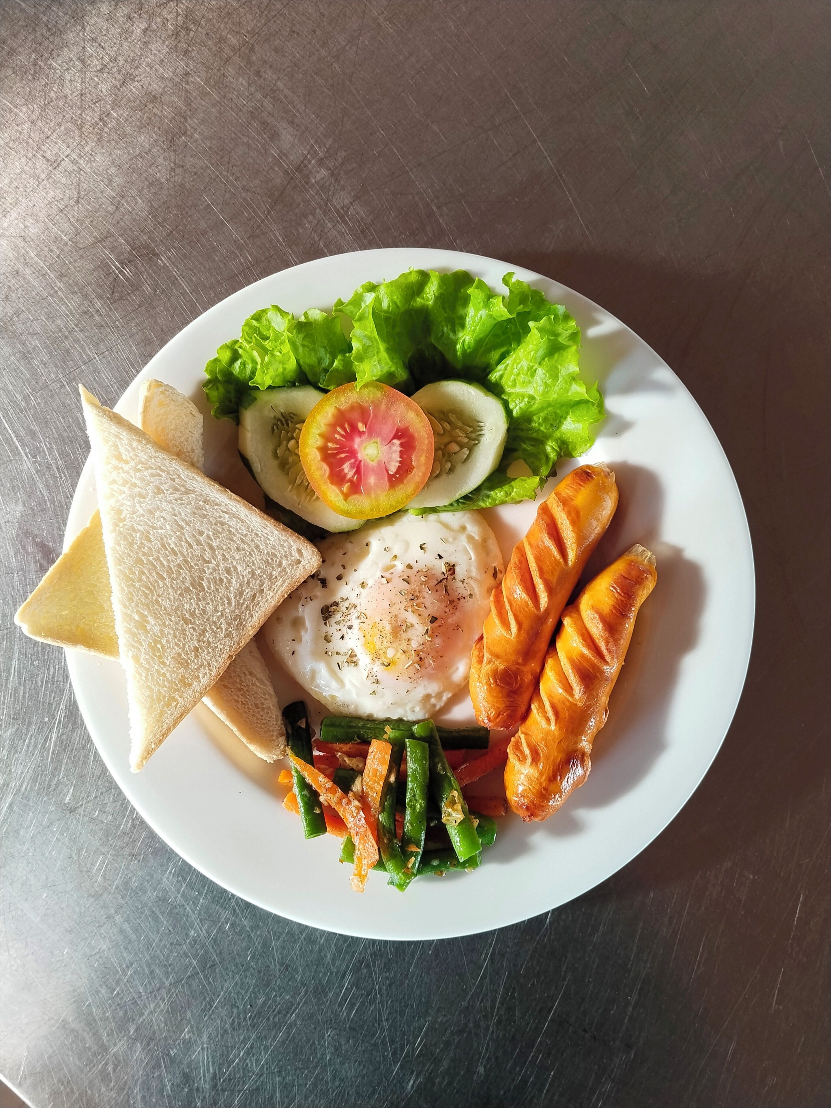 Um prato com ovo frito, torrada, alface, tomate, pepino e salsicha grelhada.