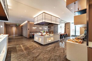 Um saguão moderno de hotel com ilha central, área de estar e uma grande janela que proporciona iluminação natural.