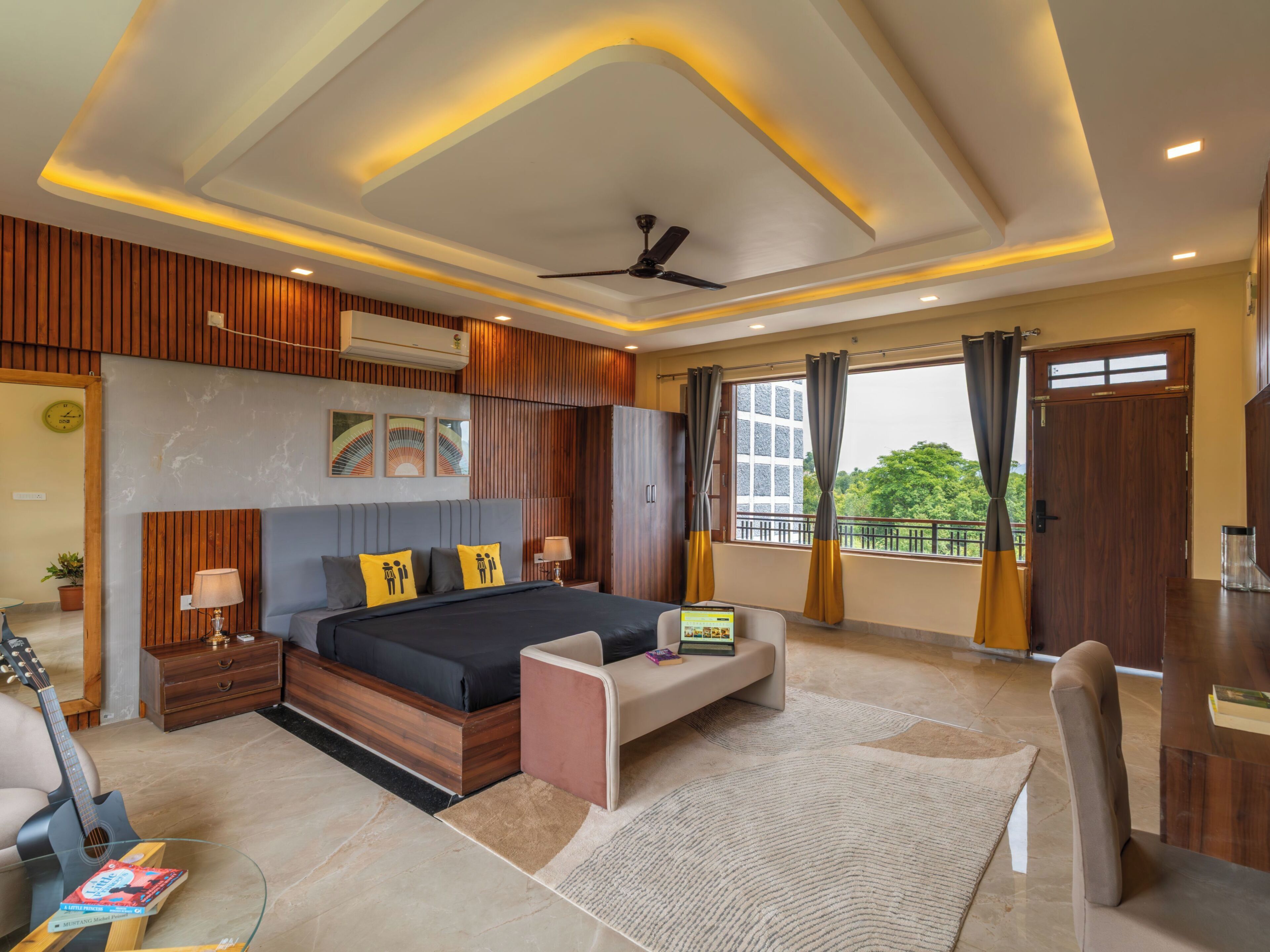 Quarto moderno com cama, mesas de cabeceira, escrivaninha, cadeira, um violão e vista para o verde lá fora.