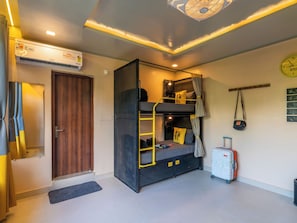 Quarto compacto com cama beliche, uma mala, relógio na parede e ventilador de teto.
