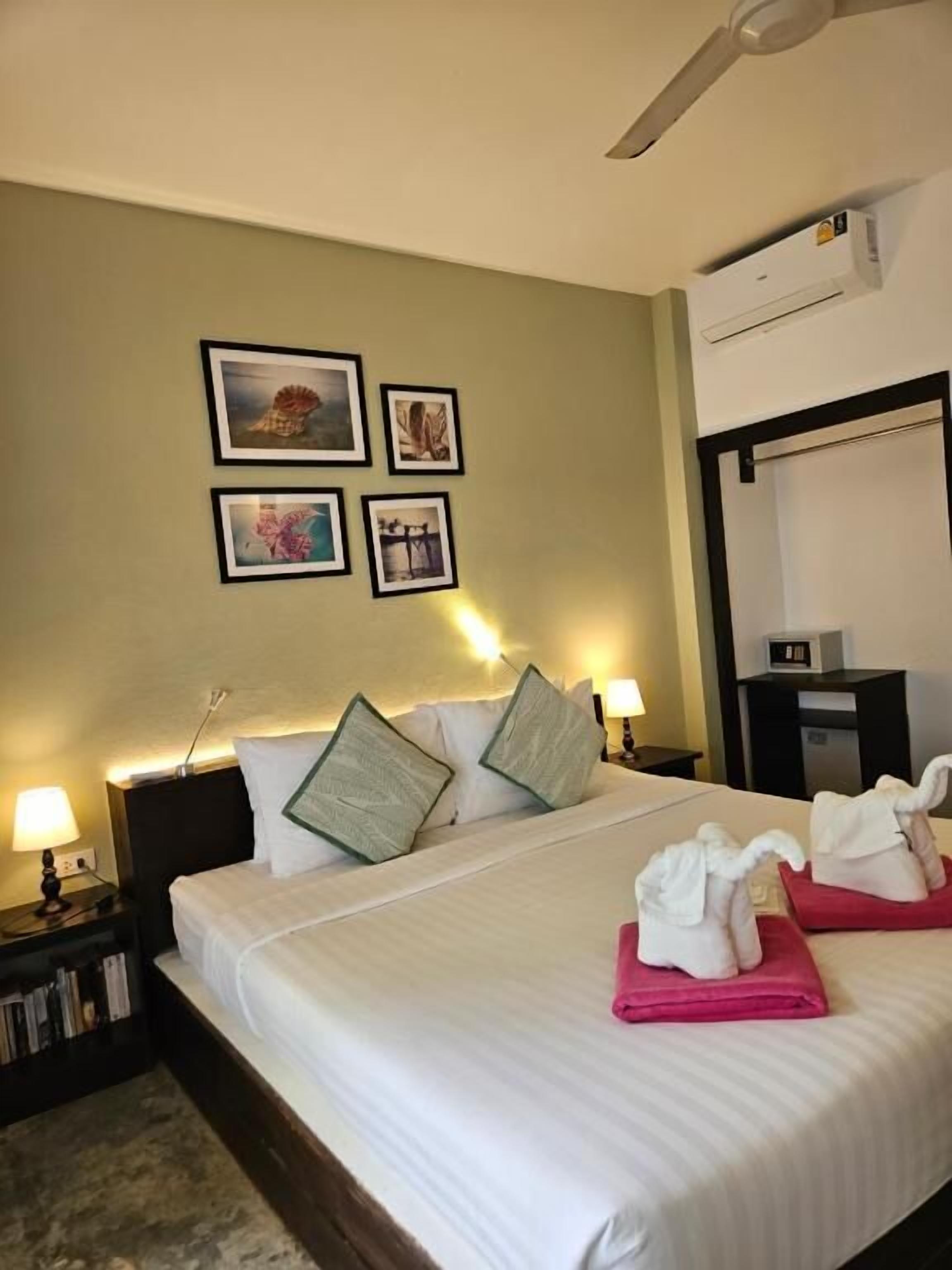 Quarto de hotel com cama, luminárias de cabeceira, ventoinha no teto, aparelho de ar condicionado e quadros nas paredes.