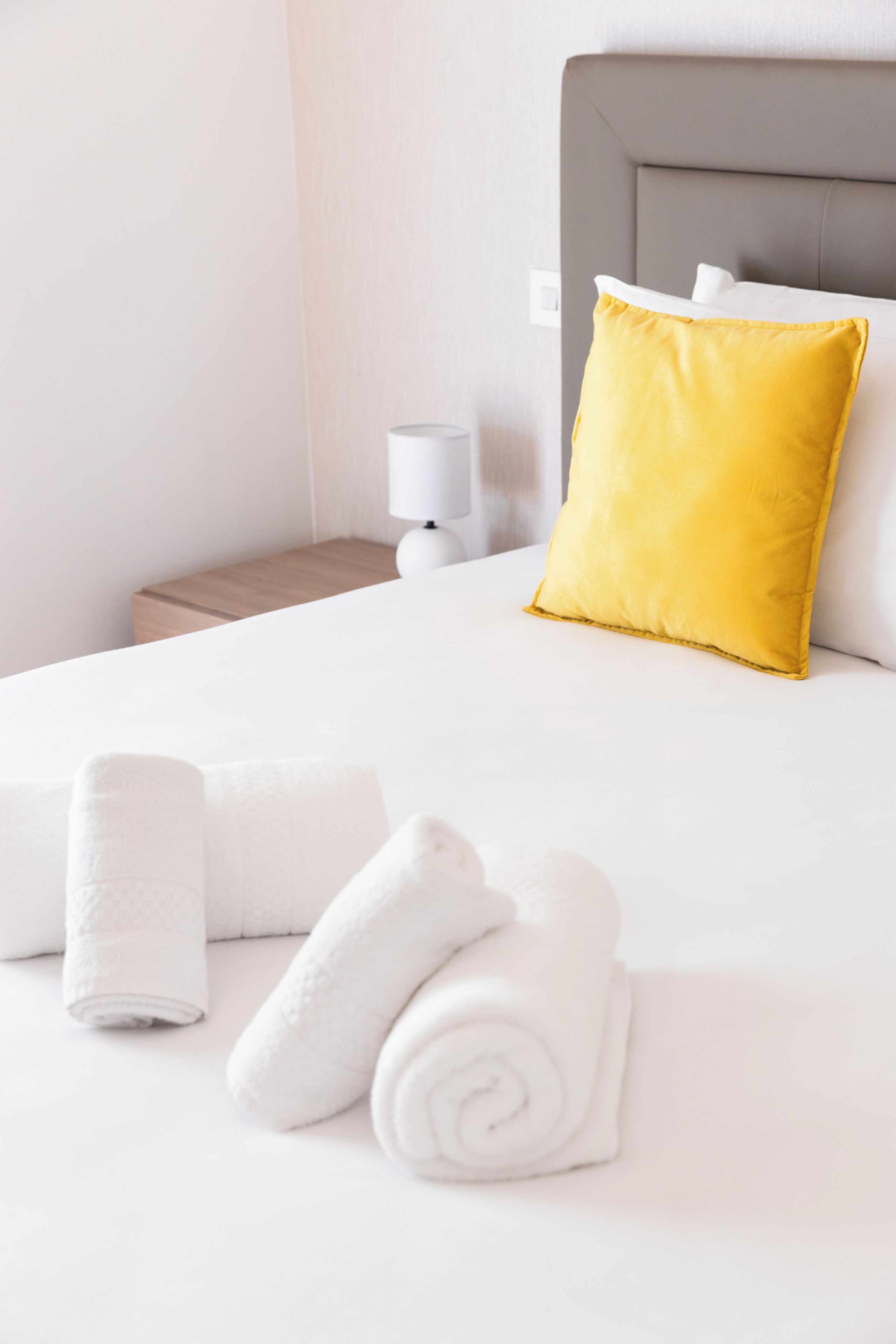 Cama bem arrumada com lençóis brancos e um travesseiro amarelo, abajur branco e mesa de cabeceira de madeira.