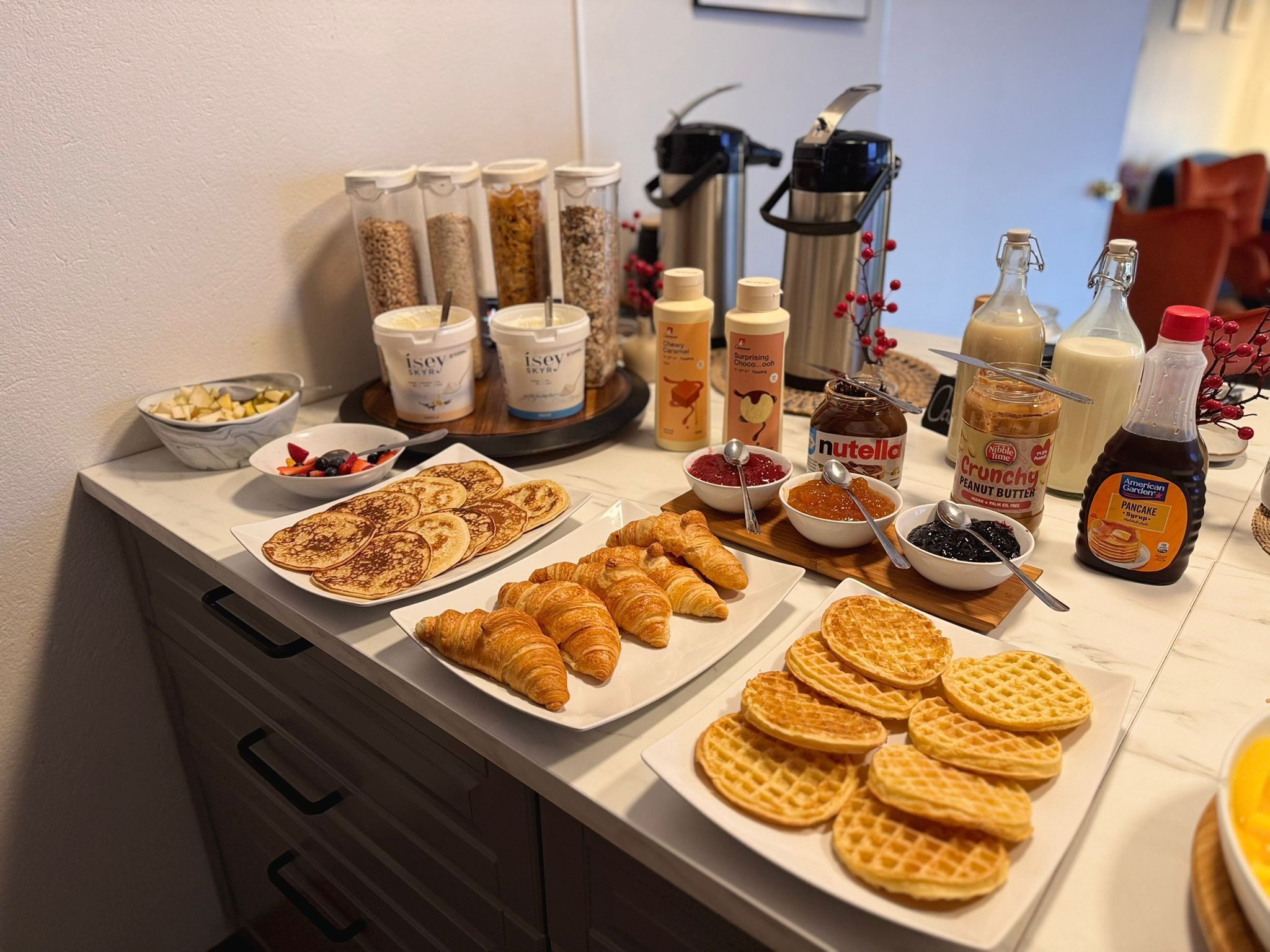 Um buffet de café da manhã com panquecas, croissants, waffles e diversas opções de acompanhamentos.