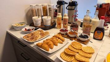 Um buffet de café da manhã com panquecas, croissants, waffles e diversas opções de acompanhamentos.