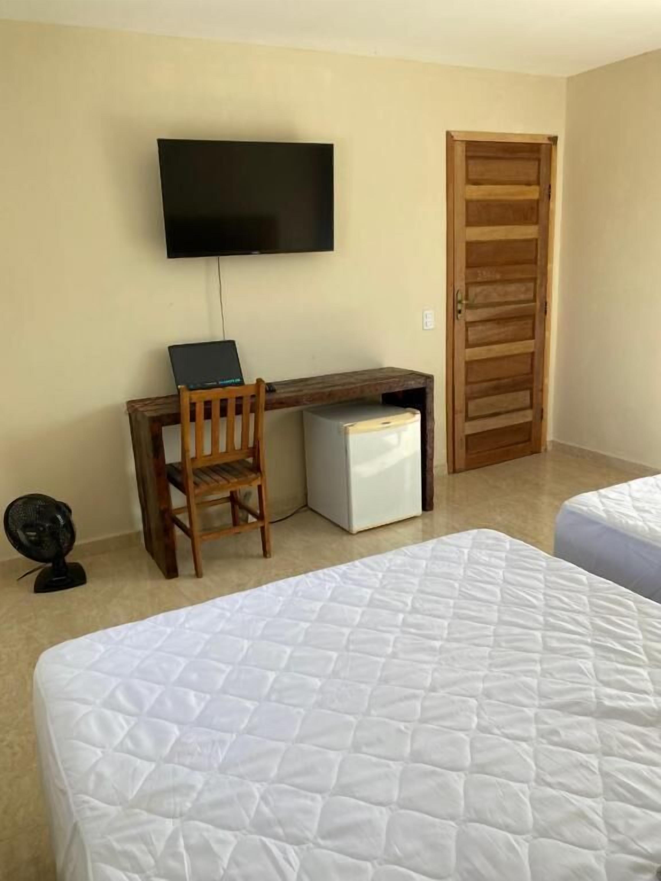 Um quarto com cama, escrivaninha com cadeira, televisão, frigorífico e porta de madeira.
