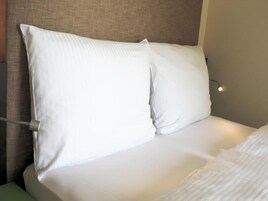 Cama bem arrumada com roupa de cama e travesseiros brancos, cabeceira e luminária de cabeceira.