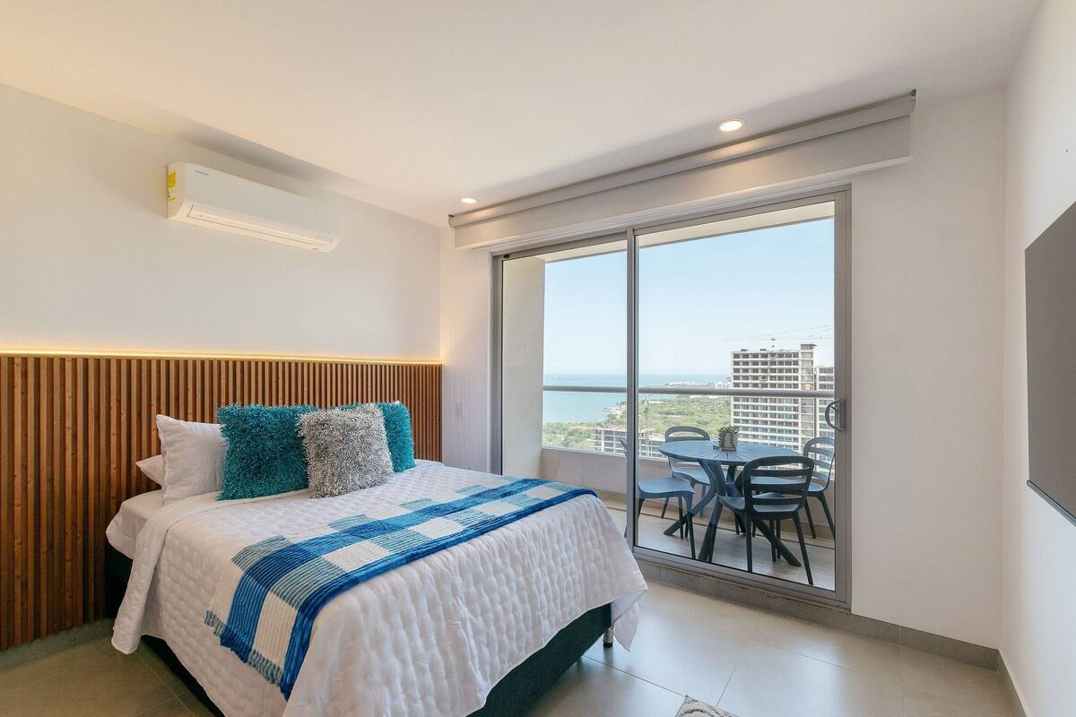 Quarto com cama, cabeceira de madeira, varanda com mesa e cadeiras e uma janela grande.