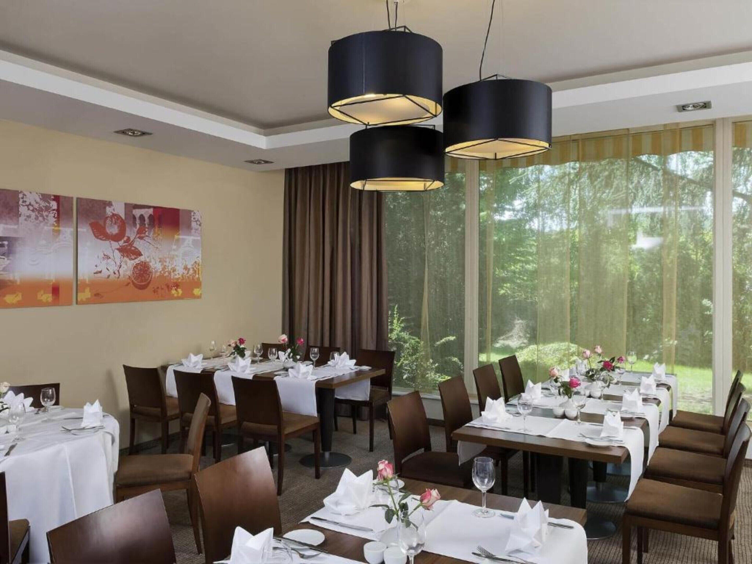 Um restaurante com várias mesas, luminárias pendentes escuras e grandes janelas com vista para o verde.