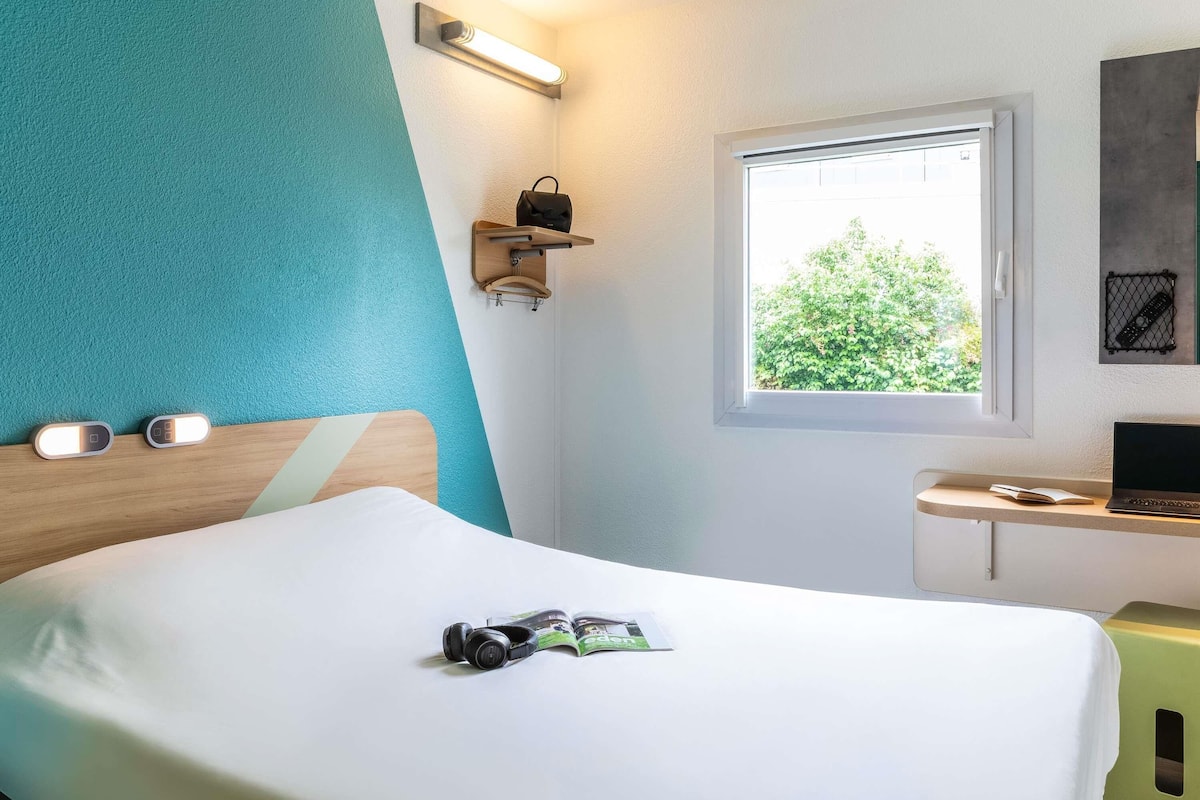 Quarto de hotel moderno com cama, mesa de cabeceira e janela com vista para o verde.