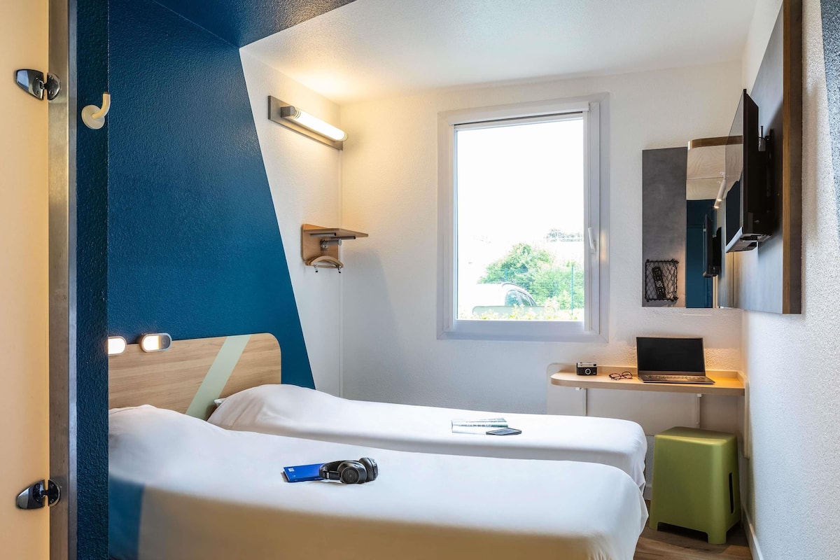 Quarto de hotel com duas camas, uma escrivaninha com um laptop, uma janela e um espelho.
