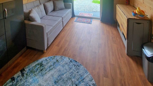 Interior acolhedor de uma cabana com um sofá, uma mesinha e vista para uma paisagem verde através de uma porta.
