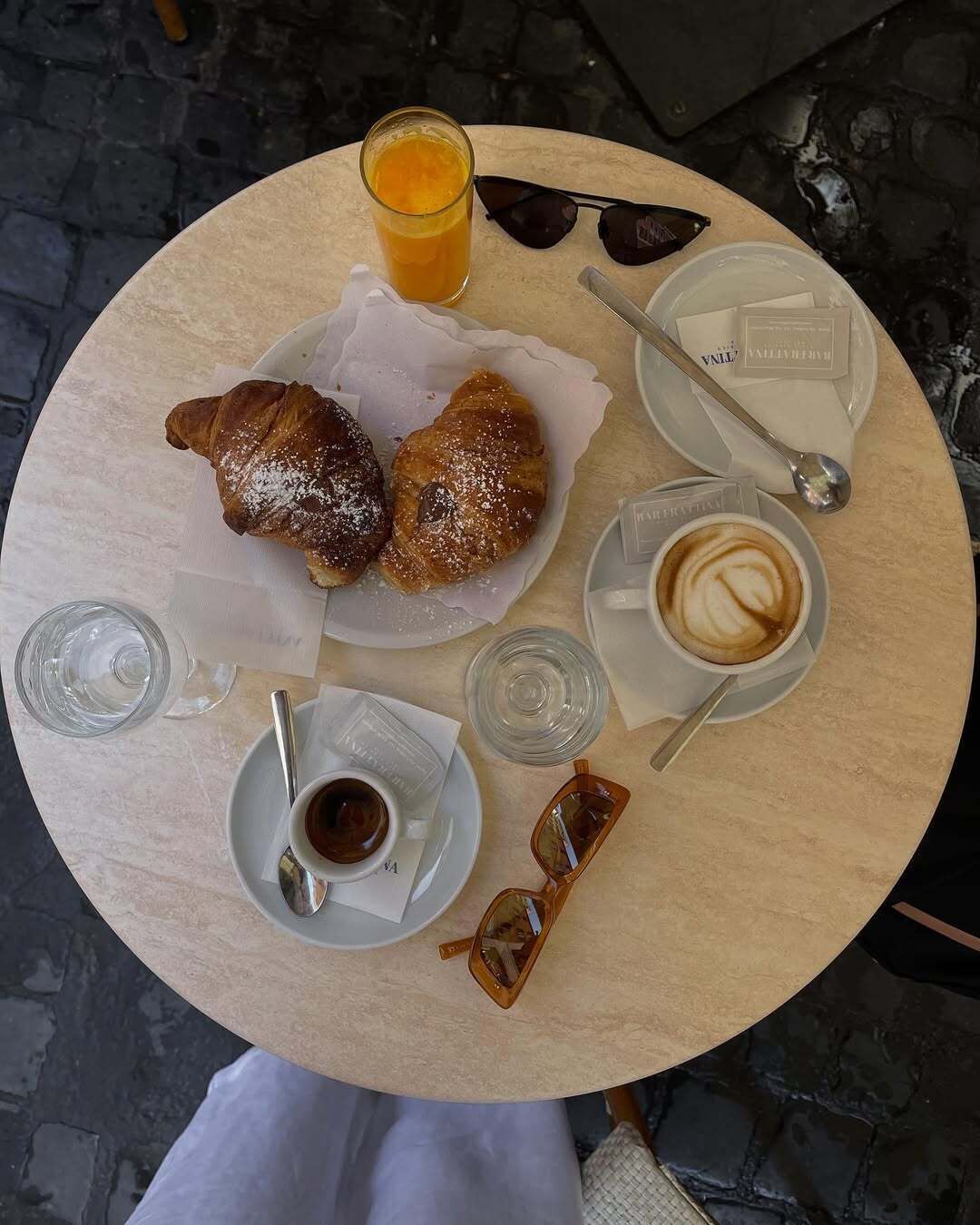 Uma mesa redonda com um croissant, um copo de sumo de laranja, uma chávena de café, óculos de sol e uma colher.
