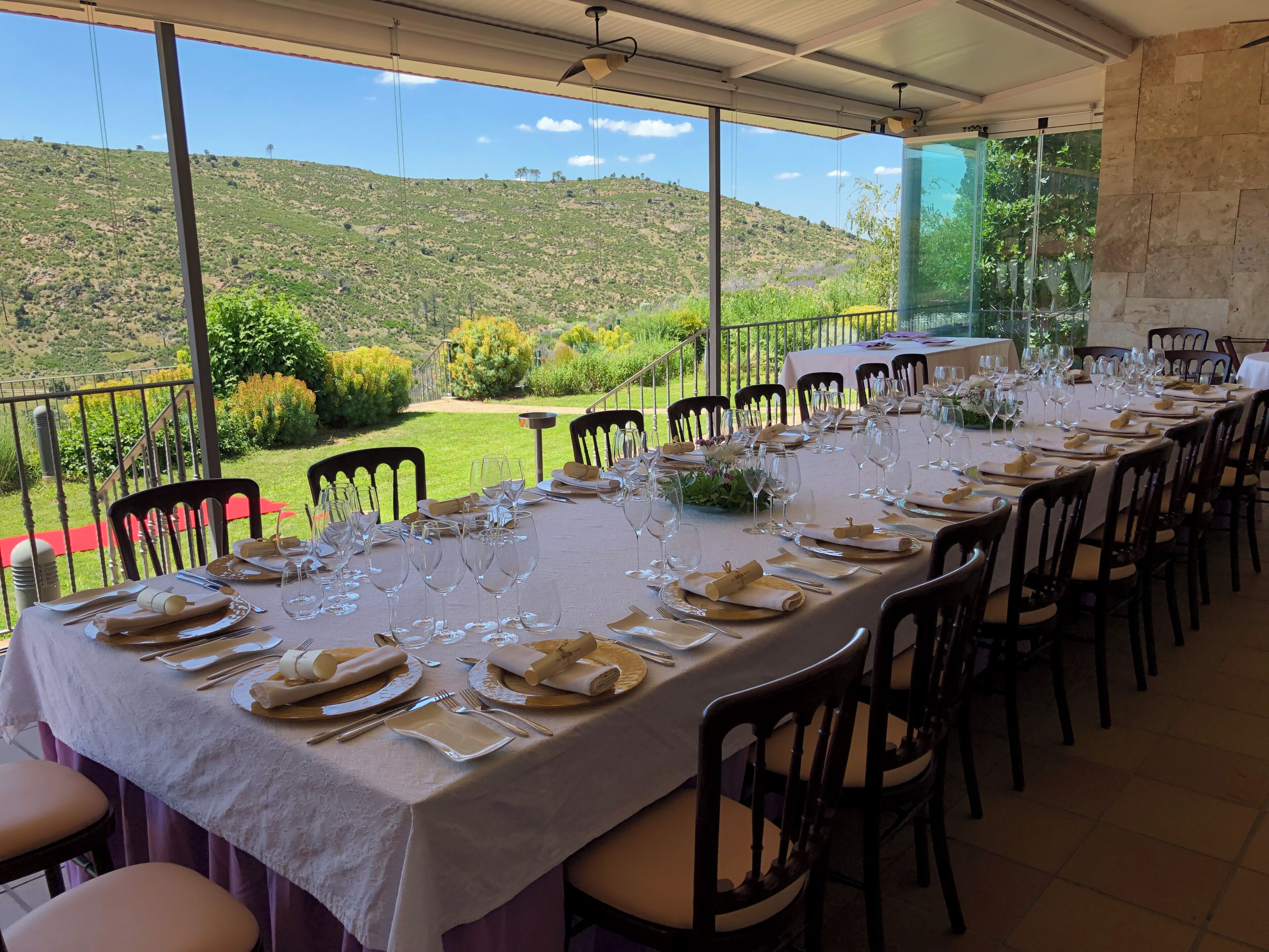 Uma longa mesa de jantar posta para uma refeição, com cadeiras, vista para uma encosta e área externa coberta.