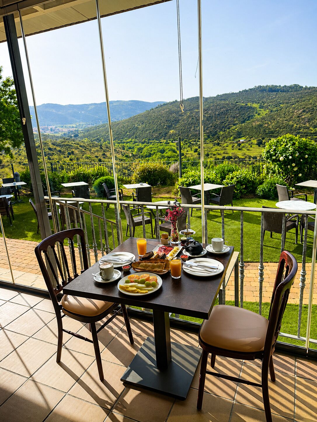 Varanda com mesa posta para café da manhã, cadeiras e vista para uma paisagem verde.