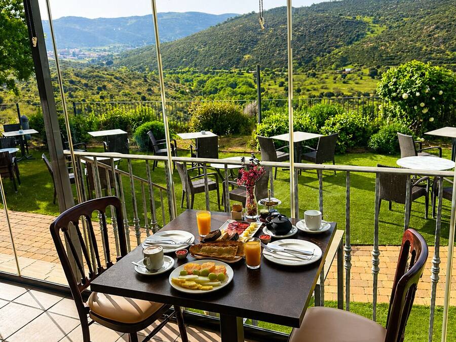 Varanda com mesa posta para café da manhã, cadeiras e vista para uma paisagem verde.