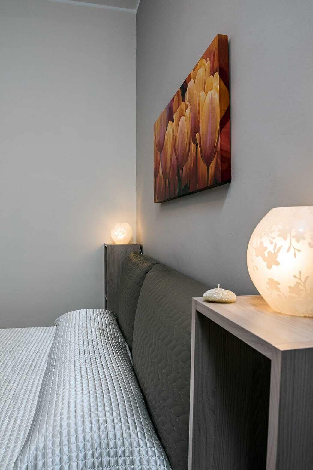 Quarto com cama, mesa de cabeceira, luminária na parede e uma pintura.