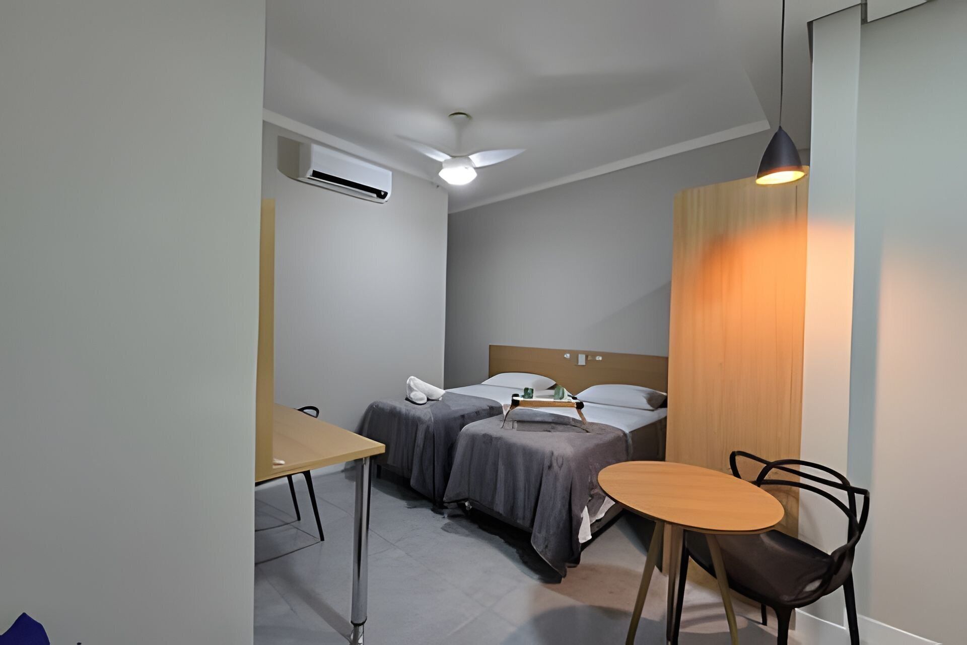 Quarto de hotel com duas camas, uma escrivaninha, uma cadeira, uma mesa, unidade de ar condicionado e ventoinha no teto.