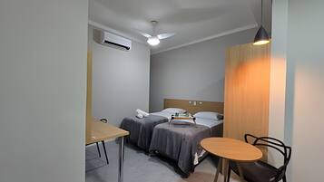 Quarto de hotel com duas camas, uma escrivaninha, uma cadeira, uma mesa, unidade de ar condicionado e ventoinha no teto.