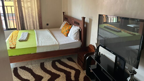 Uma cama de solteiro com cabeceira de madeira, edredom verde e branco, e um tapete estampado. Uma TV de tela plana está fixada na parede à direita.