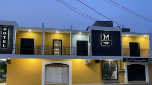 Hotel com fachada amarela, placa preta com a letra 'M' em branco e entrada de garagem visível.