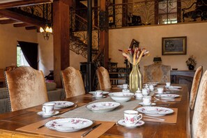 Uma sala de jantar com uma longa mesa de madeira posta para refeição, rodeada por cadeiras estofadas.