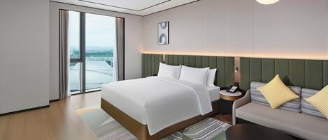 Quarto de hotel moderno com uma cama grande, um sofá, uma mesa redonda e vista da cidade através de amplas janelas.