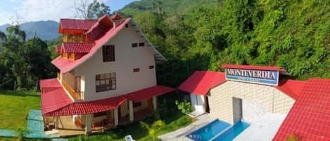 Um edifício de dois andares com telhado vermelho, piscina e uma placa com a inscrição 'Monteverde'.