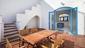Um pátio com mesa e cadeiras de madeira, uma escadaria e uma porta de vidro que dá para um quarto com um sofá azul.