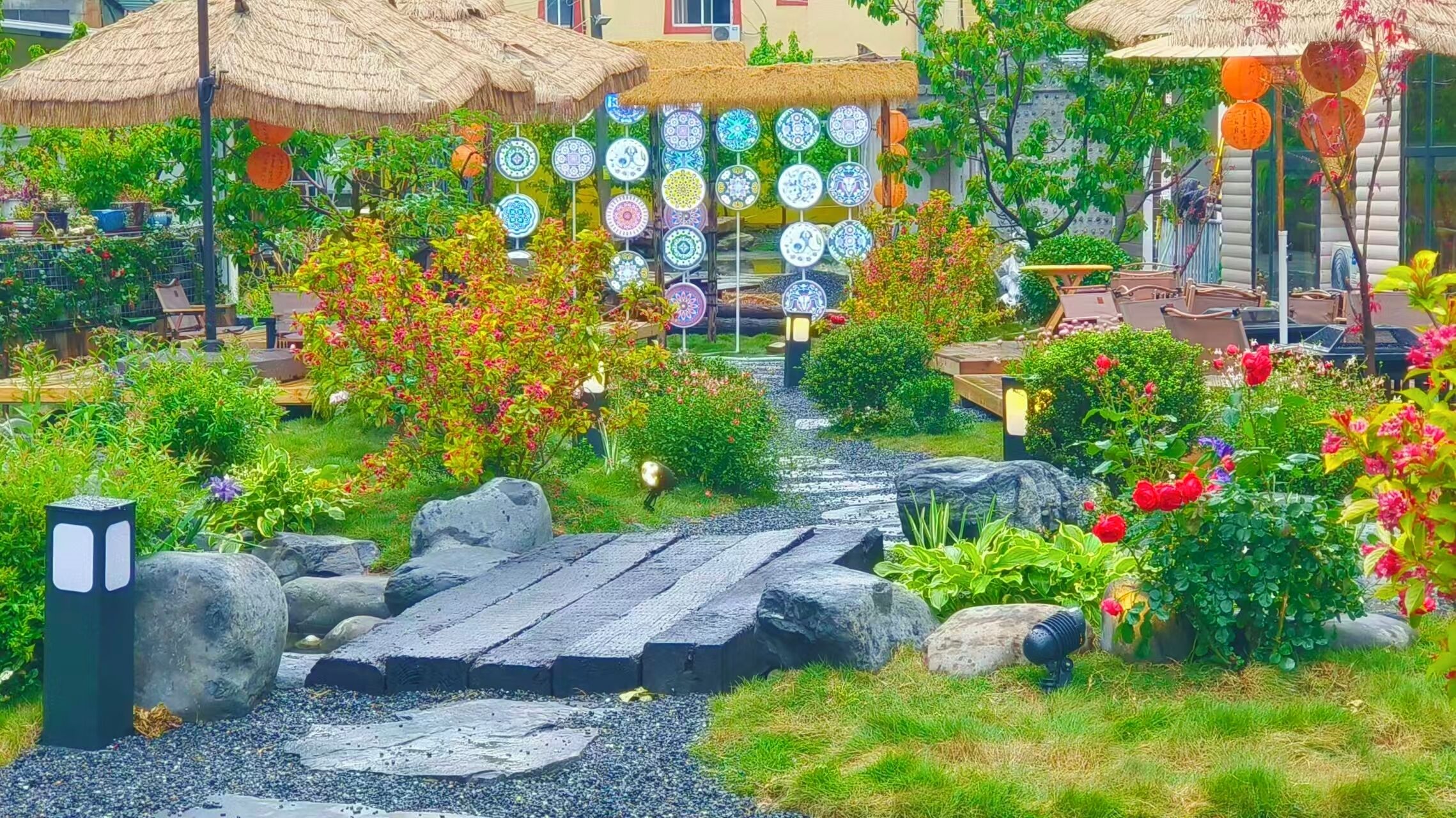 Um caminho de jardim com placas decorativas, uma construção com telhado de palha e vegetação exuberante.