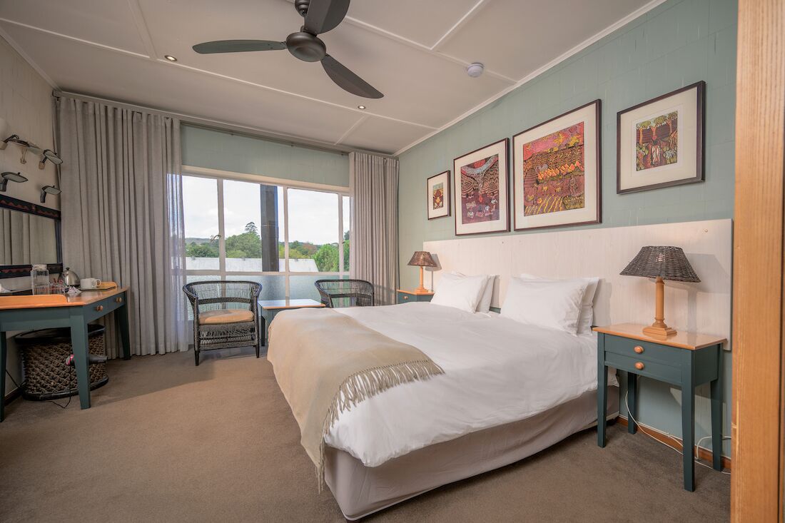 Quarto de hotel com cama, duas mesas de cabeceira, escrivaninha com cadeira, ventoinha no teto e quadros nas paredes.