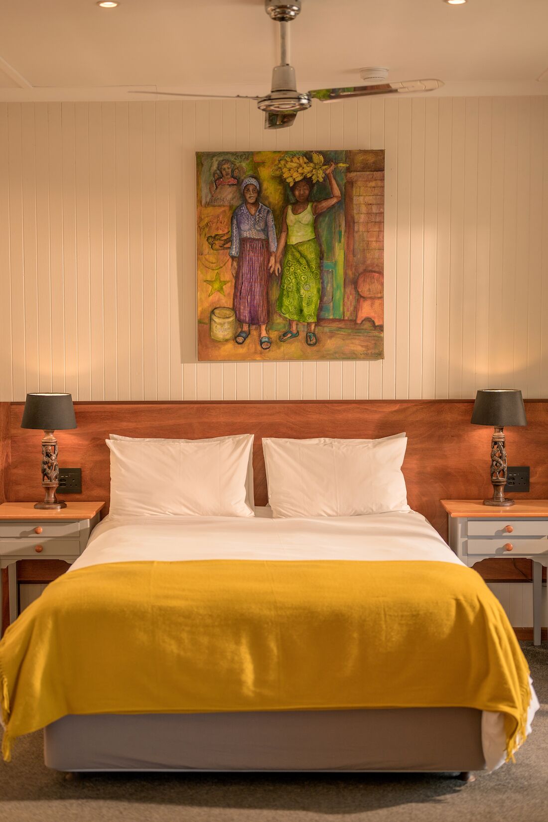 Quarto de hotel com uma cama grande, duas mesas de cabeceira com luminárias, um ventilador de teto e uma pintura na parede.