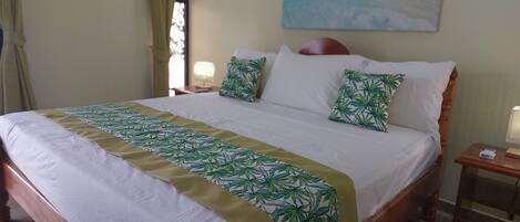 Cama com colcha estampada em folhas verdes, almofadas brancas e cabeceira de madeira.