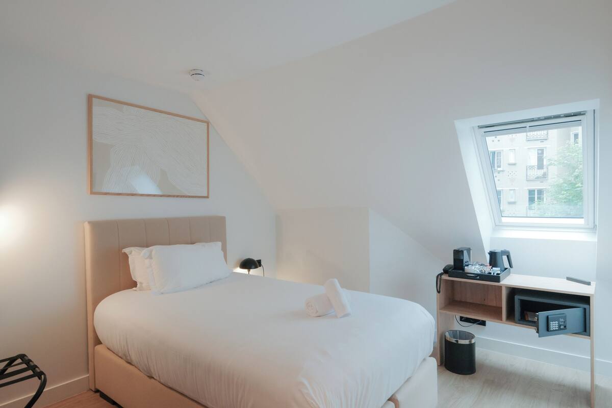 Quarto moderno com cama, mesa de cabeceira, abajur e janela com vista para edifícios.