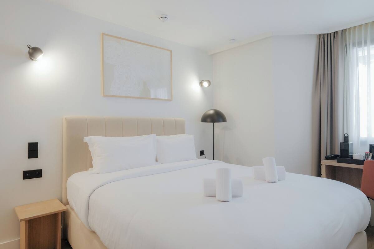 Quarto de hotel com uma cama grande, abajures, uma mesinha e uma janela com cortinas.