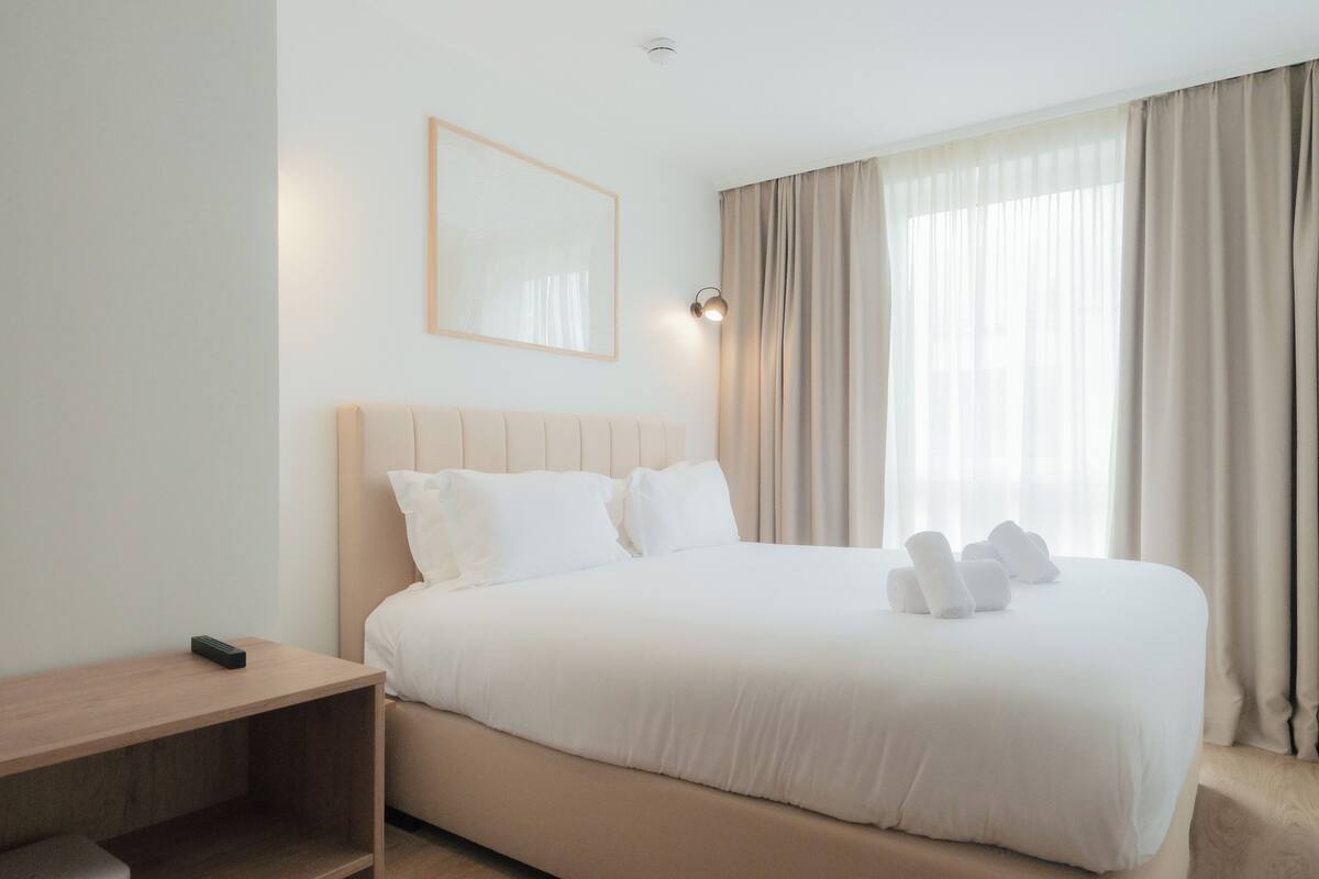 Quarto de hotel com cama, mesa de cabeceira, luminária e janela com cortinas.