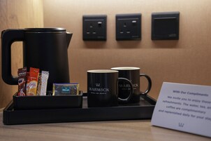 Uma estação de café com uma cafeteira de café preto, duas canecas com a inscrição 'Warwick Hotels and Resorts' e diversos pacotes de café.