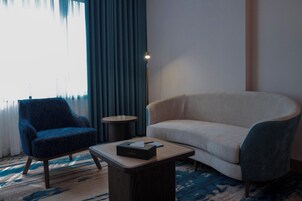 Um quarto com uma poltrona azul, um sofá bege, uma mesa redonda pequena e um abajur.