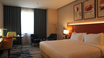 Quarto de hotel com uma cama, duas poltronas, uma escrivaninha, um abajur e uma janela com cortinas.