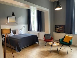Quarto de hotel com cama, escrivaninha, duas cadeiras, abajur e um quadro na parede.