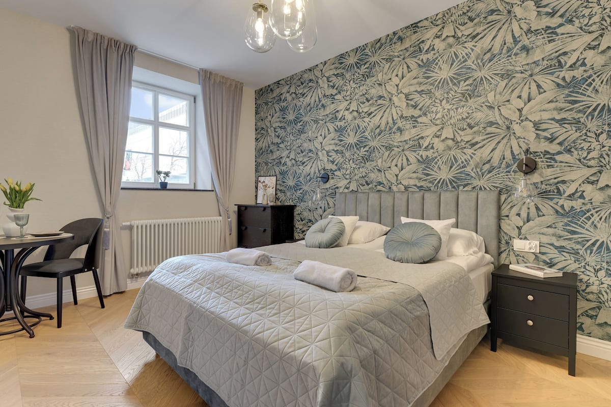 Quarto com cama, mesa de cabeceira e janela com cortinas.
