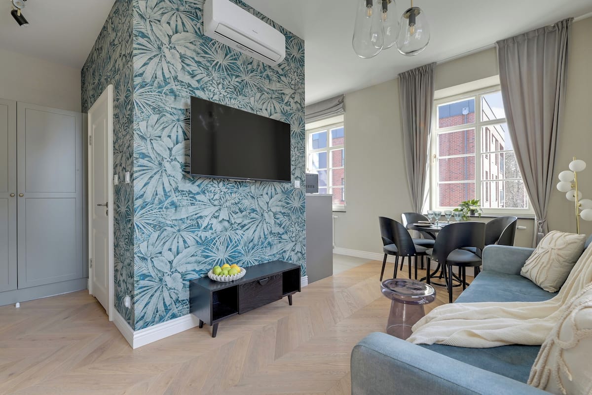 Sala de estar com televisão de tela plana fixada em parede estampada, sofá cinza, área de jantar com mesa e cadeiras, e janela com cortinas.