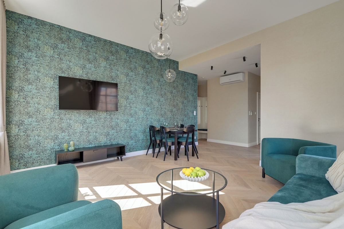 Uma sala de estar moderna com sofás verde-água, uma mesa de centro de vidro com frutas, uma área de jantar com mesa e cadeiras, e uma TV fixada numa parede estampada.