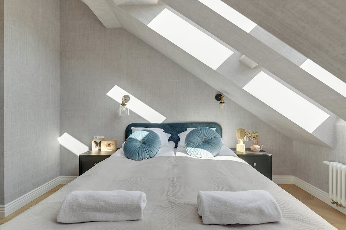 Quarto com teto inclinado, claraboias, cama com roupa de cama branca, duas almofadas, mesa de cabeceira com abajur e cômoda.