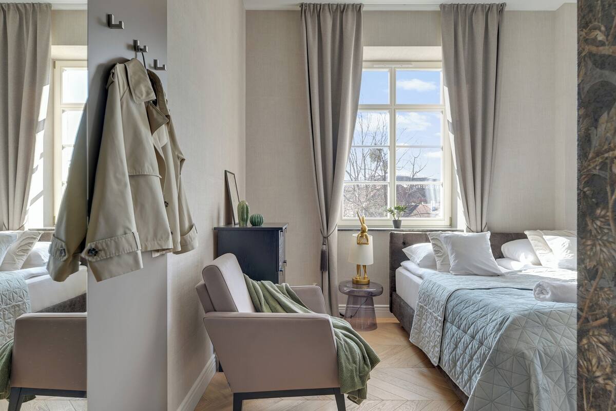 Quarto de hotel com duas camas, uma cadeira, uma mesinha com abajur e uma janela com cortinas.