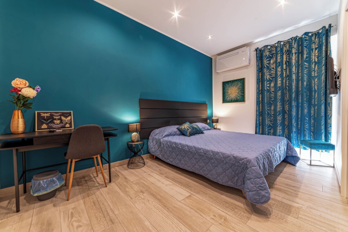 Quarto com cama, escrivaninha, cadeira e pequena mesa com um vaso de flores.