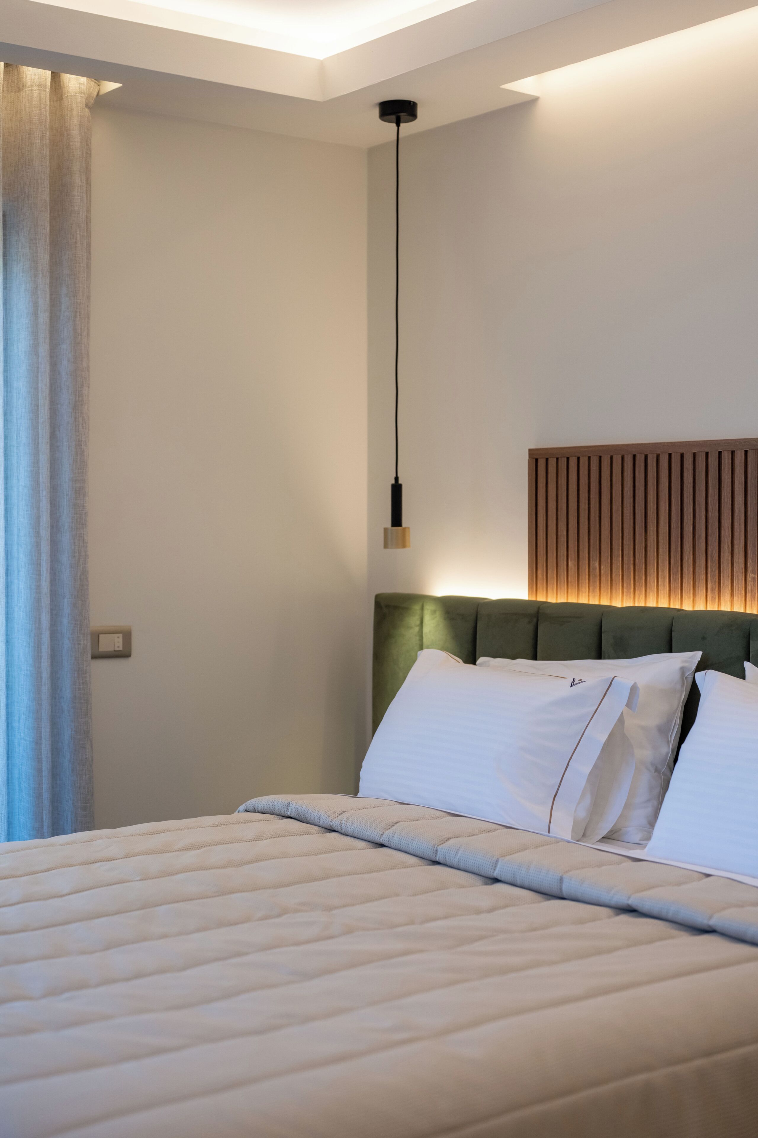 Cama bem arrumada com cabeceira, luminária pendente e janela com cortinas.