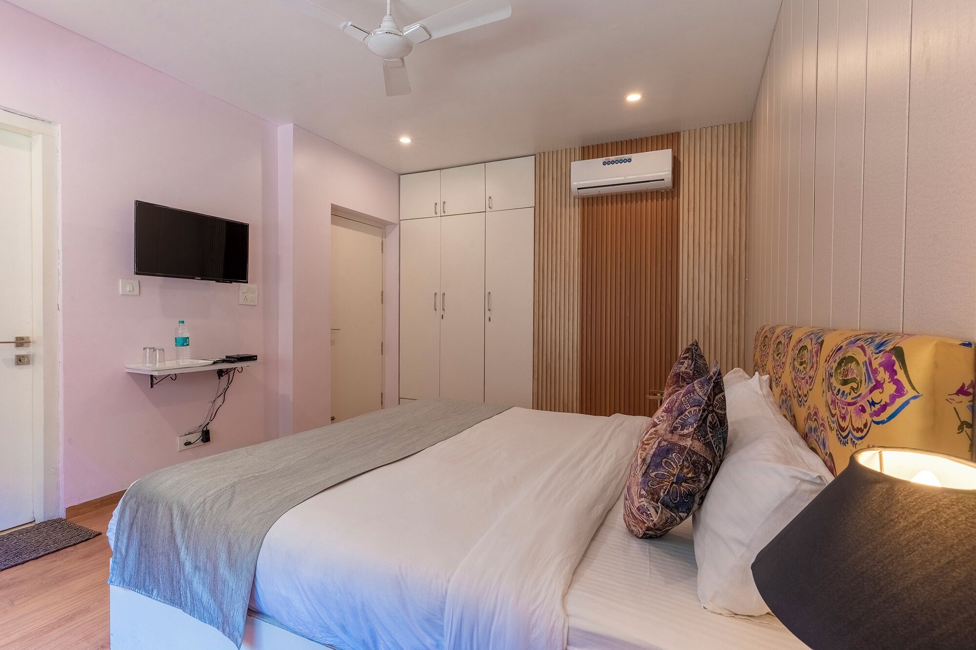 Quarto com cama, televisão, ar condicionado e mesa de cabeceira com uma garrafa e copos.